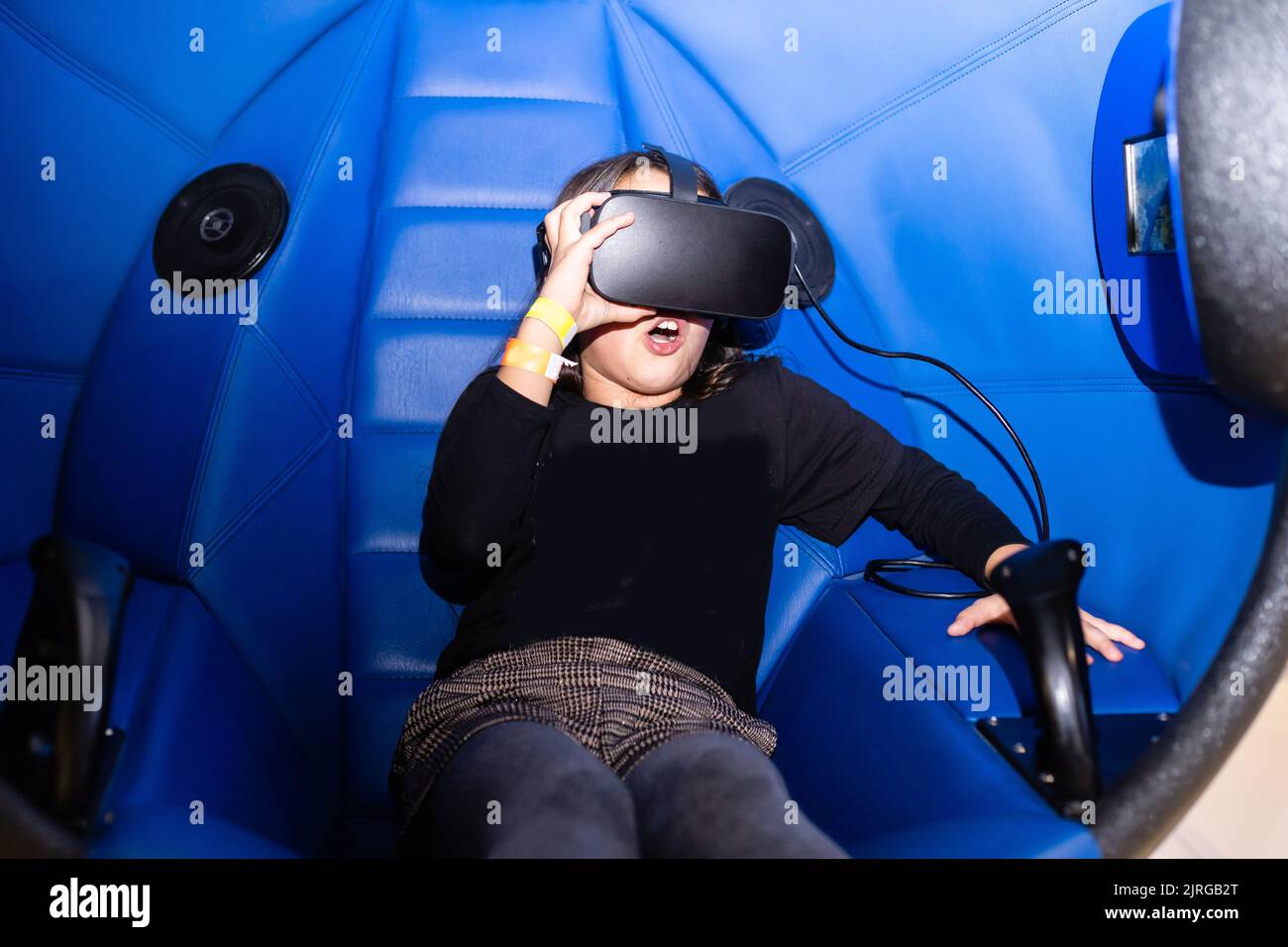 A little girl sitting in a gaming cockpit and holding the race steering ...