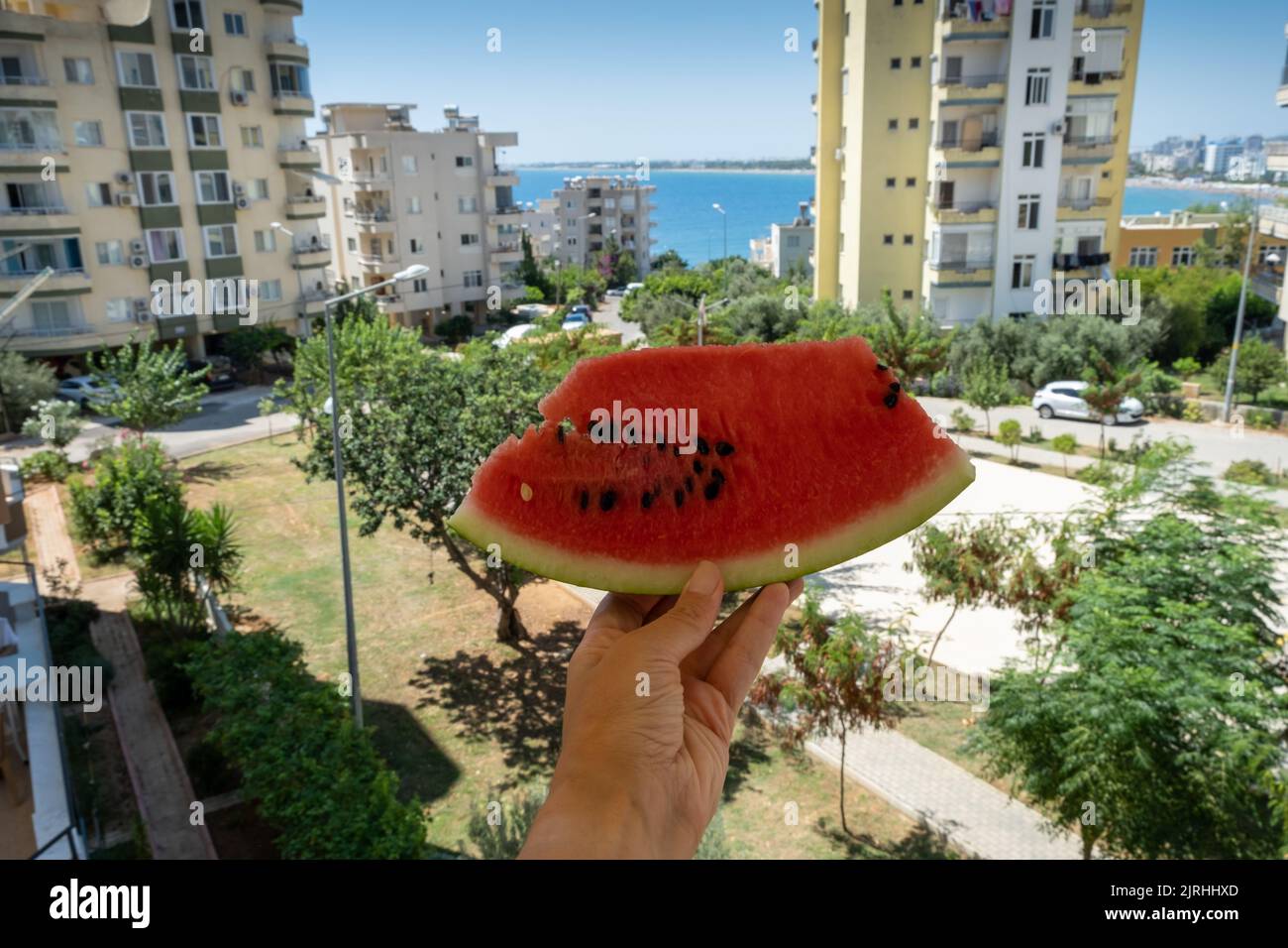 Hand holds piece of watermelon against background of beach town and sea ...