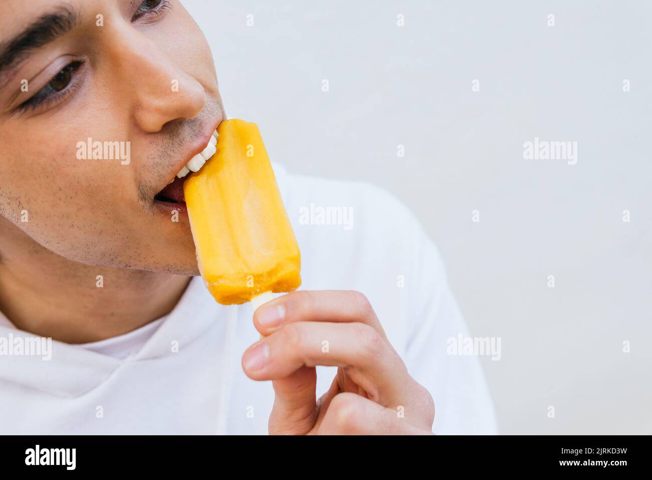 Crop happy male eating sweet orange ice lolly enjoying taste on white ...