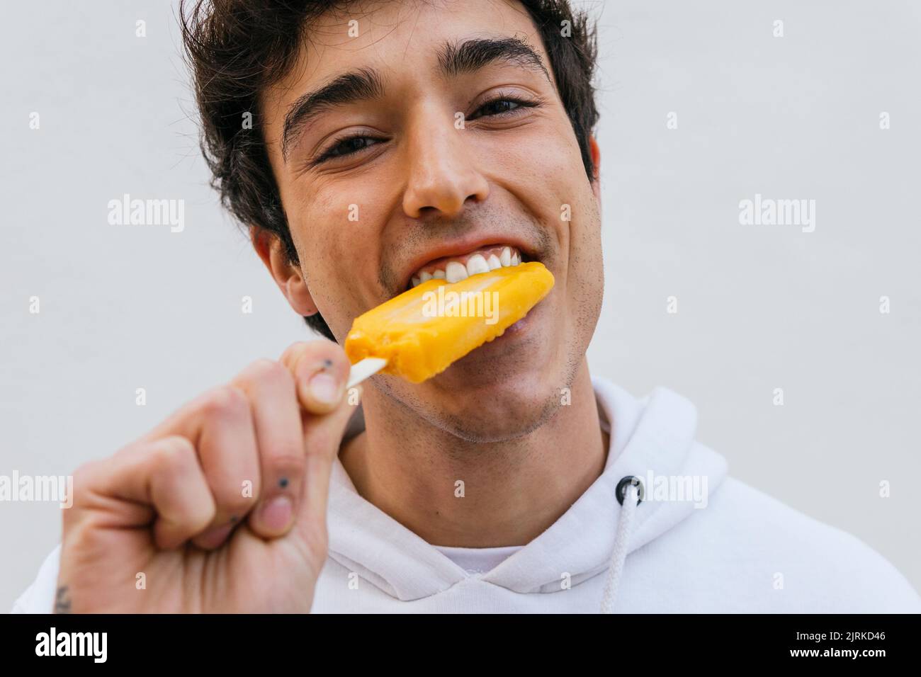 Happy young male eating sweet orange ice lolly enjoying taste on white ...
