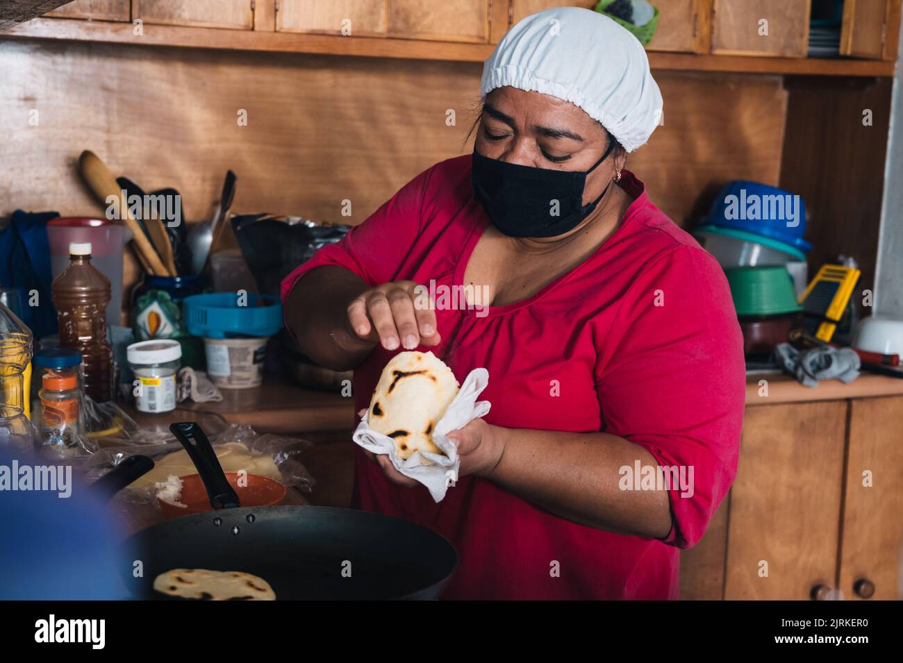 Mature ethnic female in cloth face mask cooking arepas with filling in ...