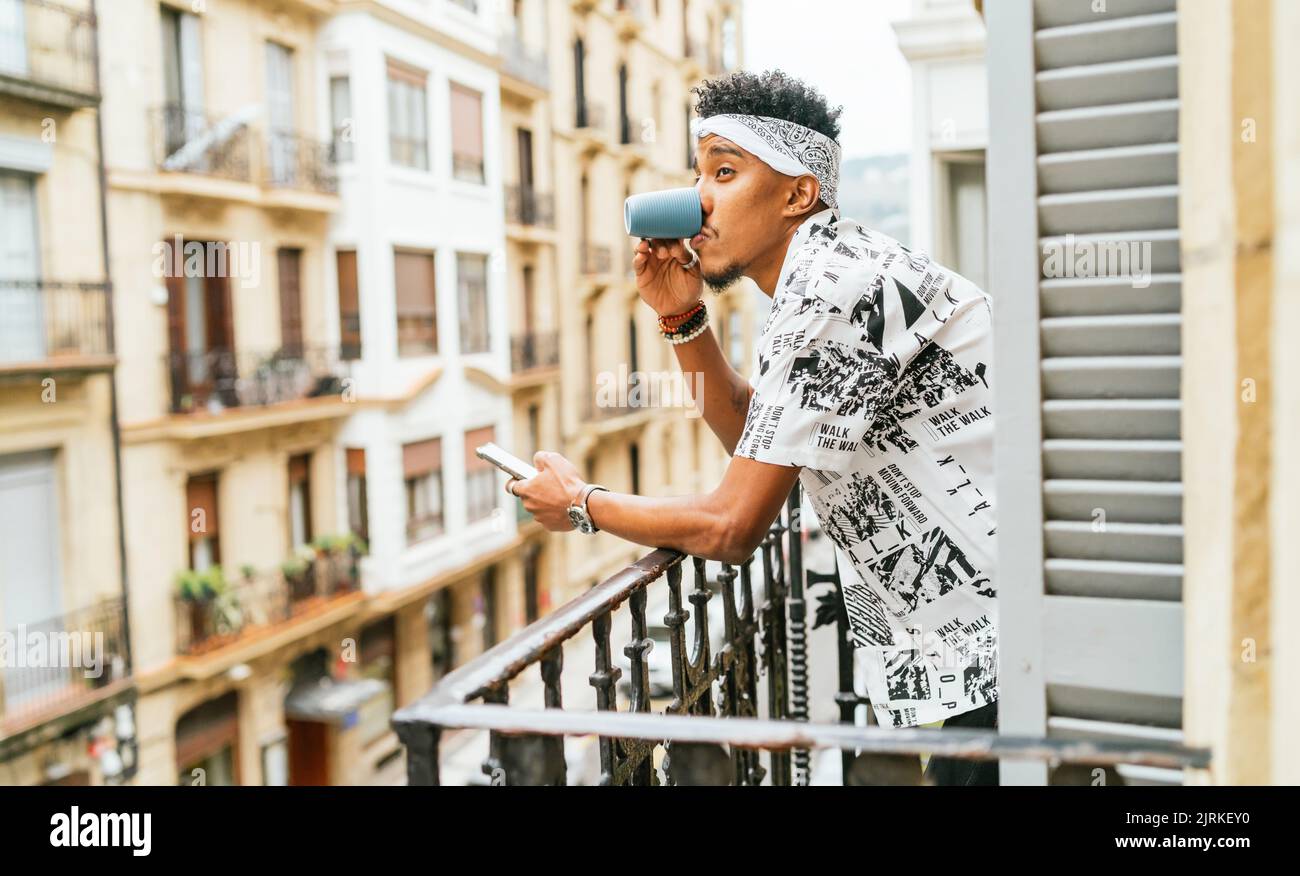Side view of cheerful ethnic male standing on balcony and browsing ...