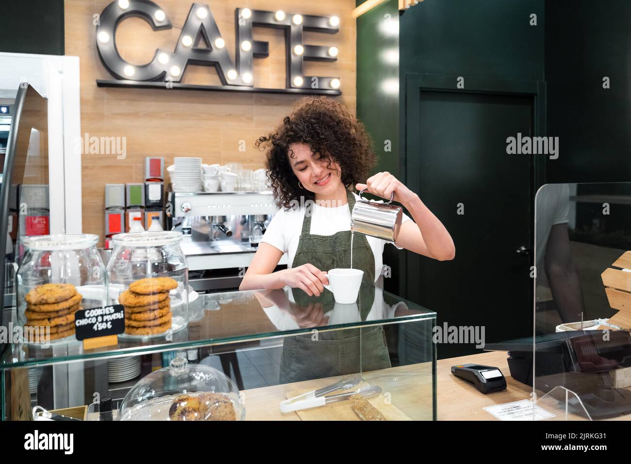 Delighted ethnic female barista in apron standing at counter in cafe ...