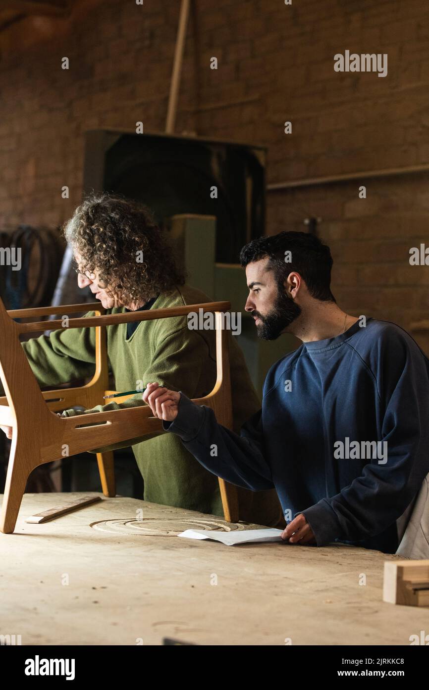 Woodworker measuring chair detail with tape while coworker writing ...