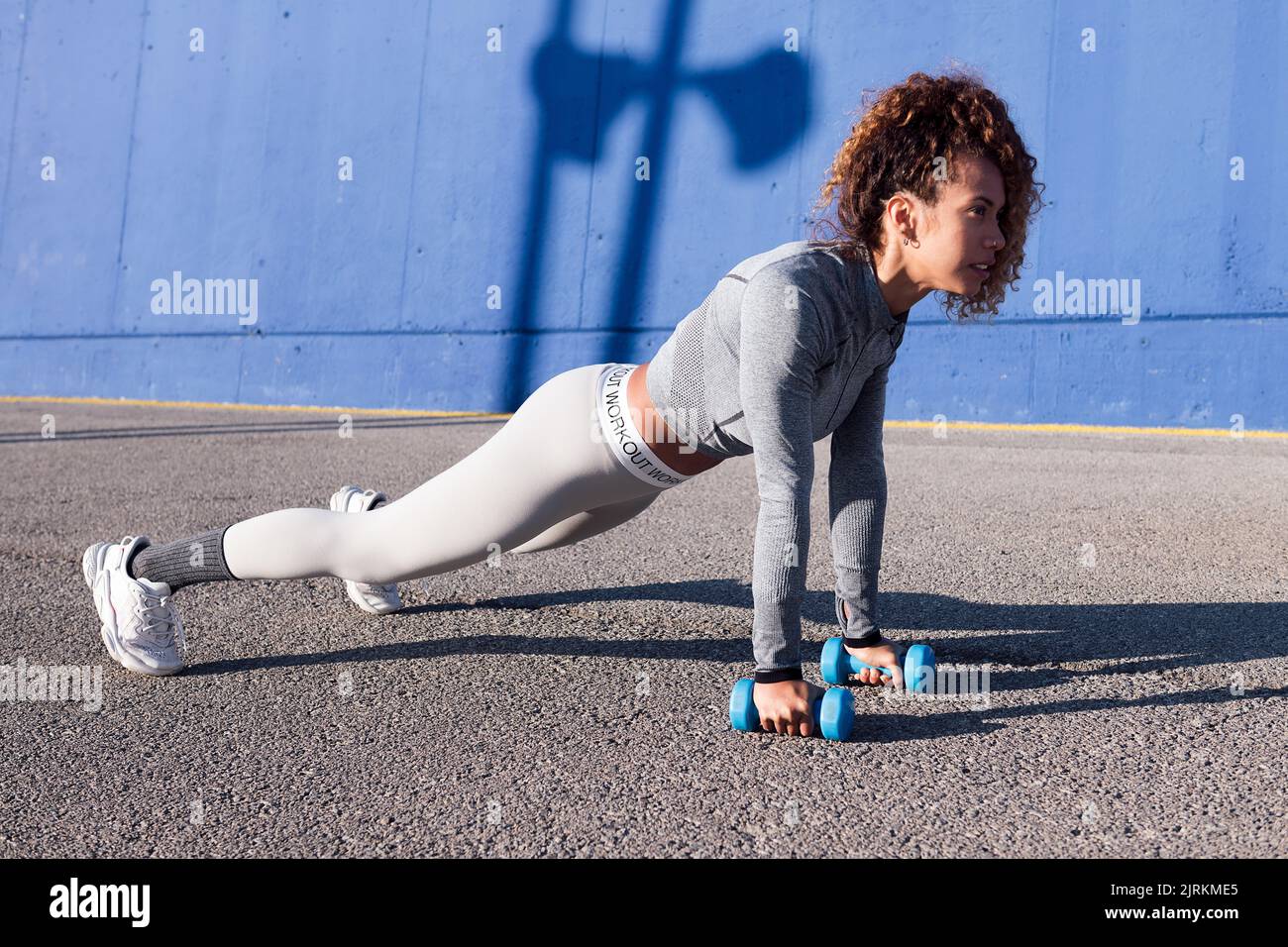 Side view of fit female athlete standing in plank pose with dumbbells ...