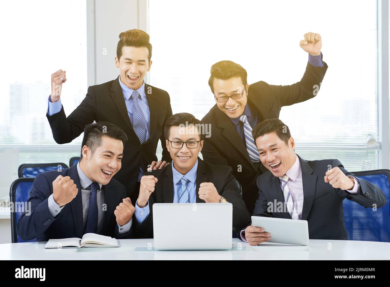 Cheerful Asian employees wearing classical suits gathered together in front of laptop and ...