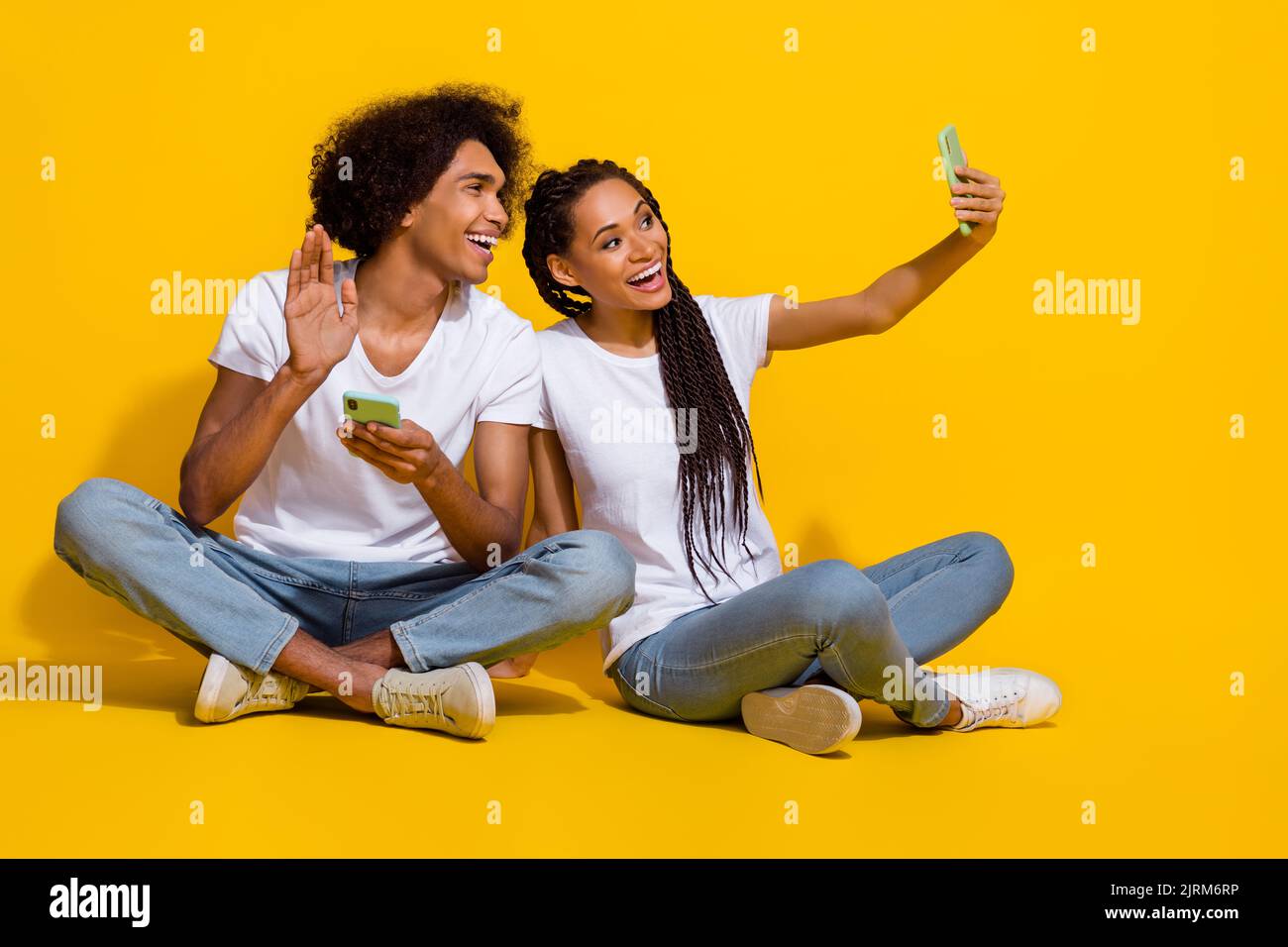 Full size photo of two cheerful people sit floor make selfie arm waving ...
