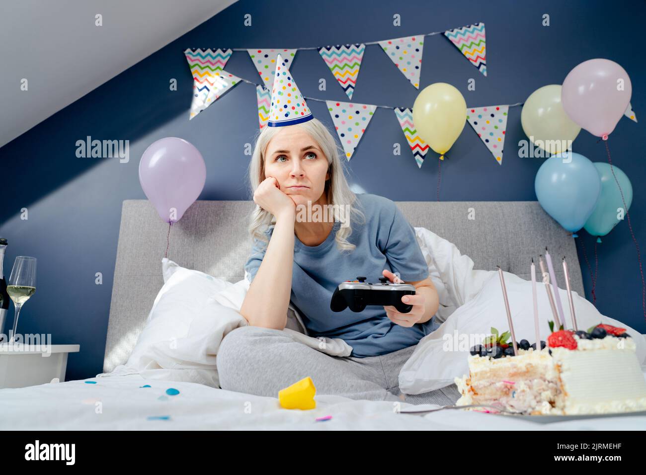 Sad, bored woman in pajamas and party cap playing video games with ...
