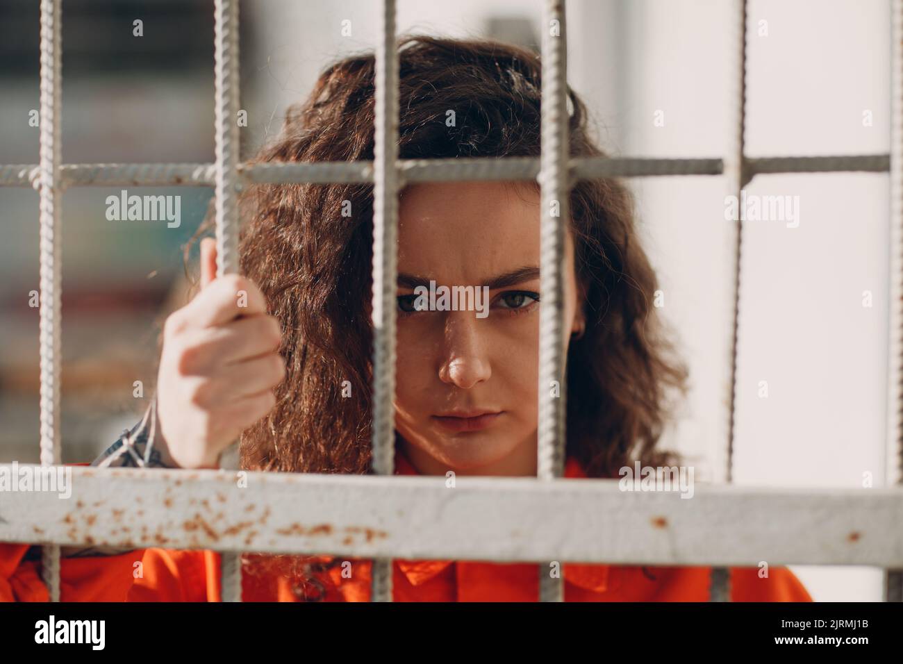Young Brunette Curly Woman In Orange Suit Behind Jail Bars Female In Colorful Overalls Portrait
