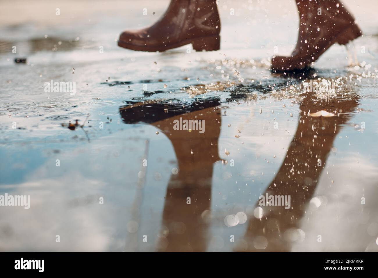 Woman wearing rain rubber boots walking running and jumping into puddle with water splash and ...