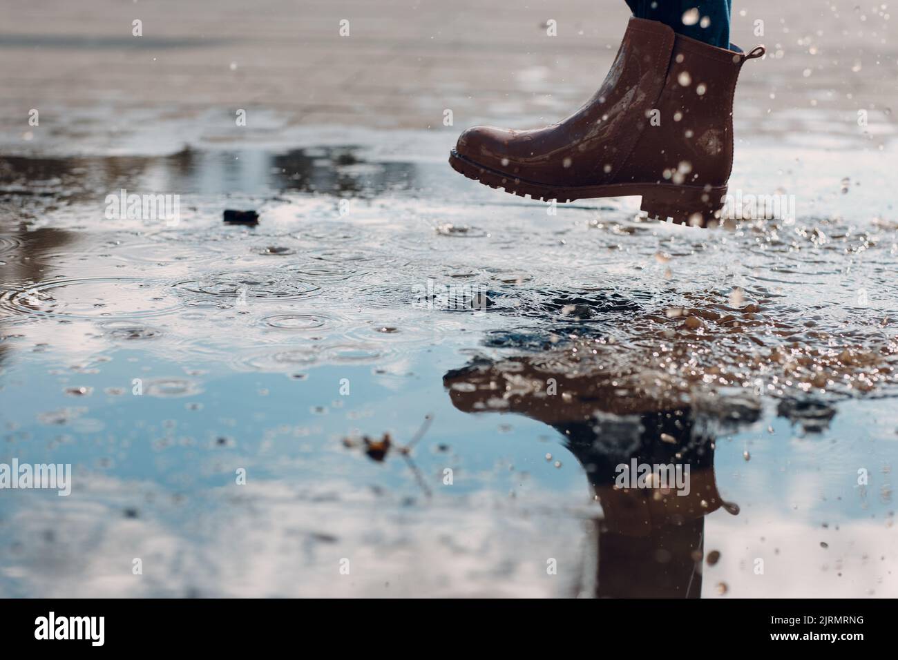 Woman wearing rain rubber boots walking running and jumping into puddle with water splash and ...