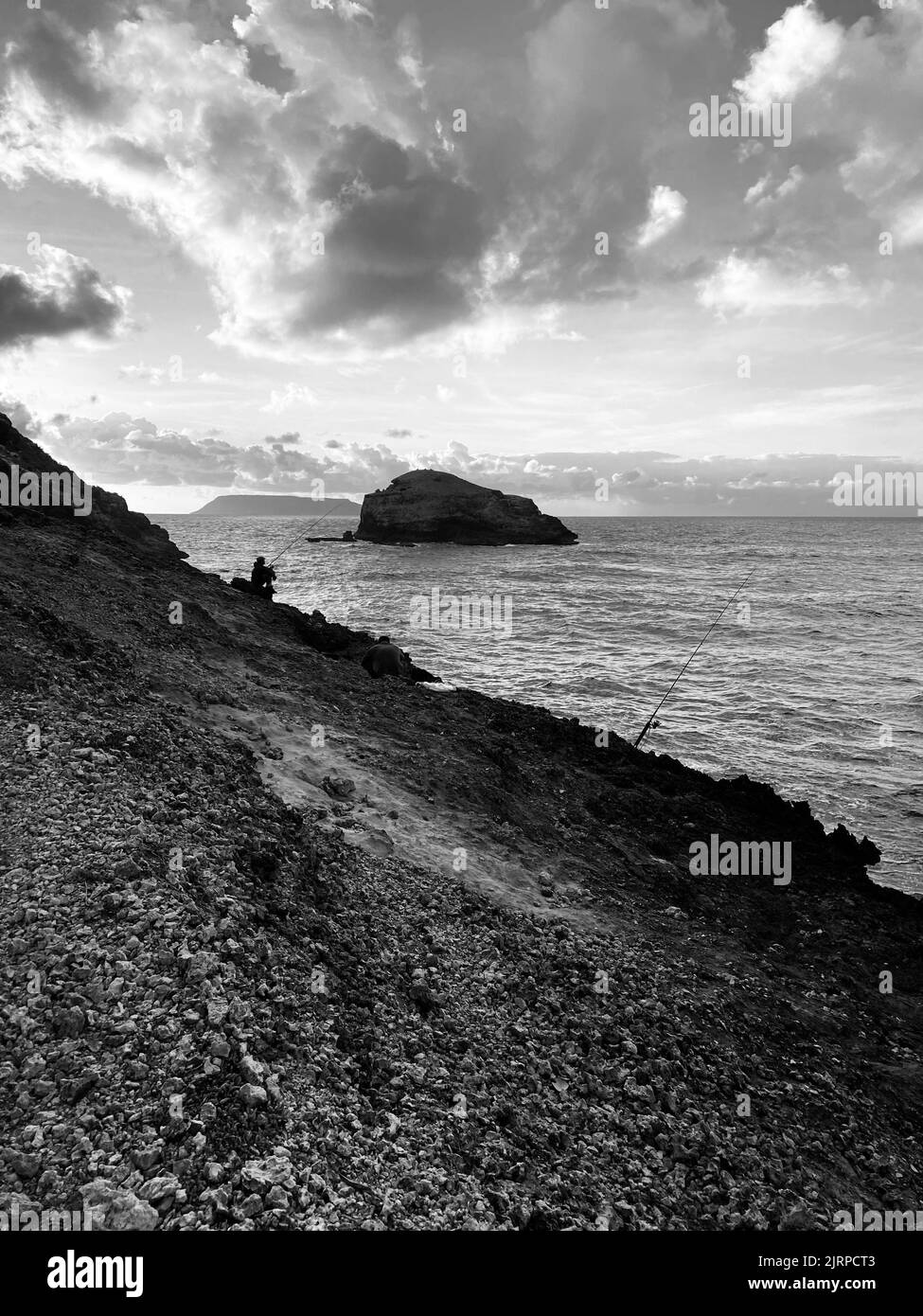 A grayscale of a beach with a huge rock in a sea under the cloudy sky ...