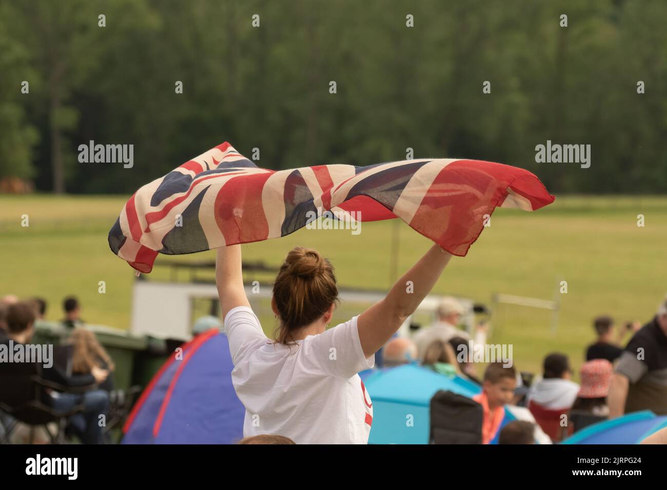 A young lady waving Union Jack flag above her head in blurred crowd ...