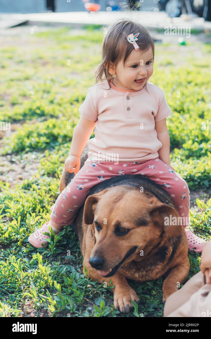 Front view of a baby girl riding her dog during a summer day in the ...