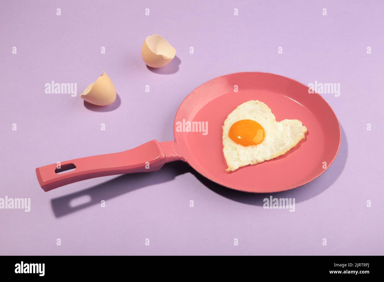 Creative idea with a pink frying pan and egg in heart shape on white and pink background ...