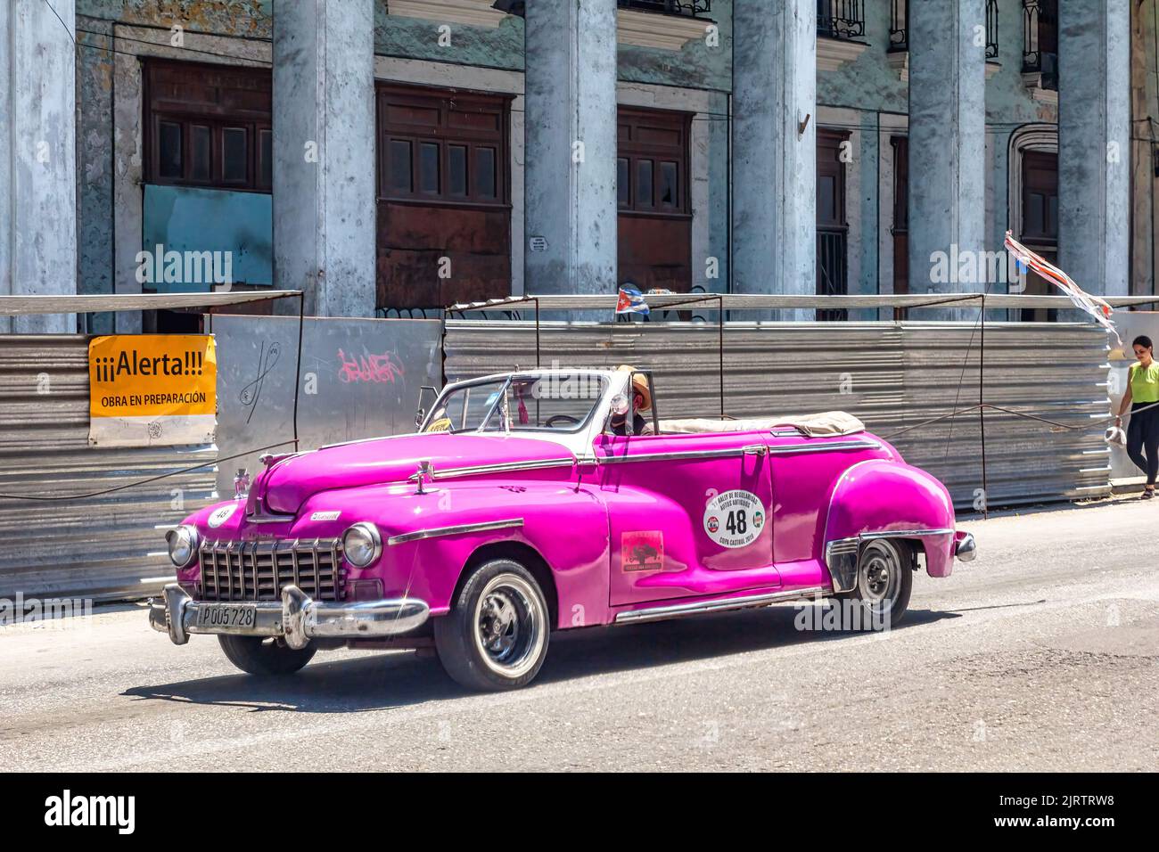 An old American convertible car drives by a collapsed building which is ...