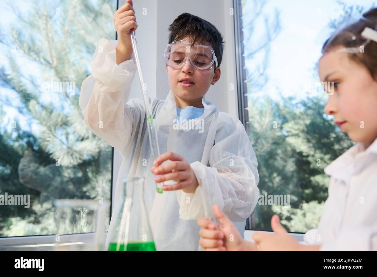 Handsome schoolchild in lab safety wear using pipette, dripping reagent ...