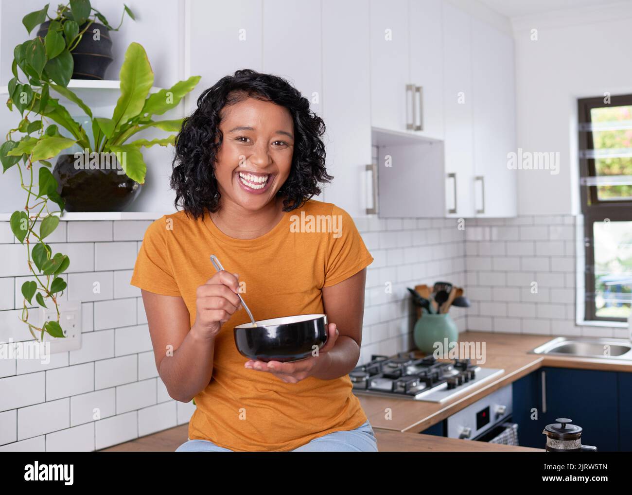 A young multi-ethnic woman sits on her kitchen counter eating breakfast ...