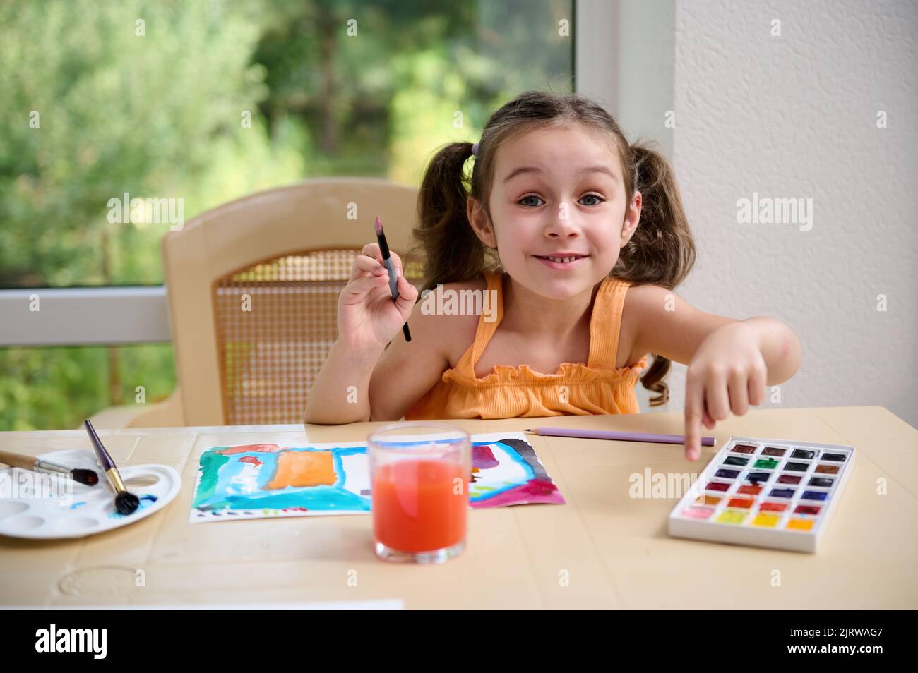 Happy little girl sitting at a table and cutely smiling at the camera ...