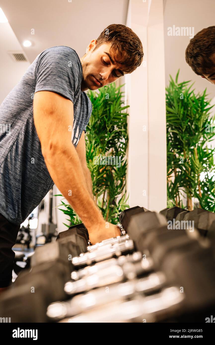 Low angle of powerful sportsman taking dumbbells from rack and checking arm muscles during ...