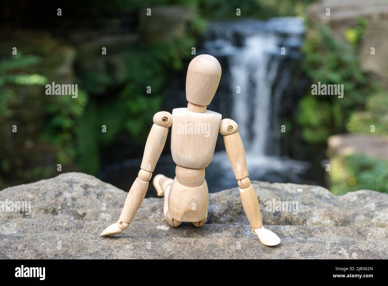 A wooden artist's figure sitting on a rock looking at a waterfall ...