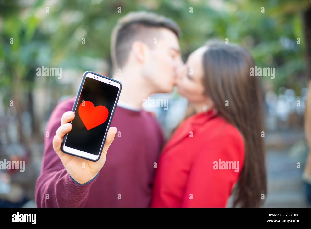 Young couple kissing while showing a smartphone with an heart shape in ...