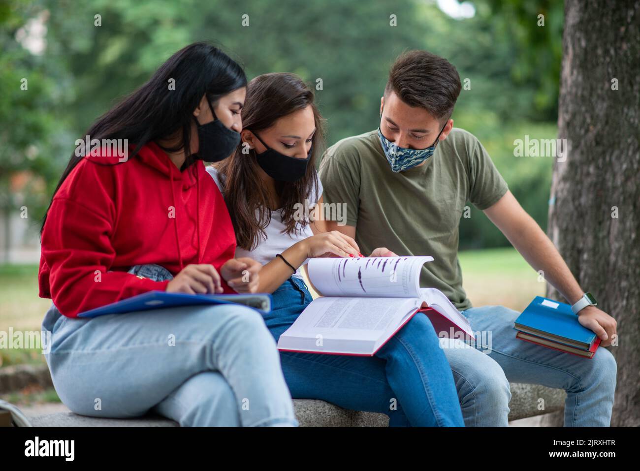 Three students studying together sitting on a bench outdoor and wearing ...