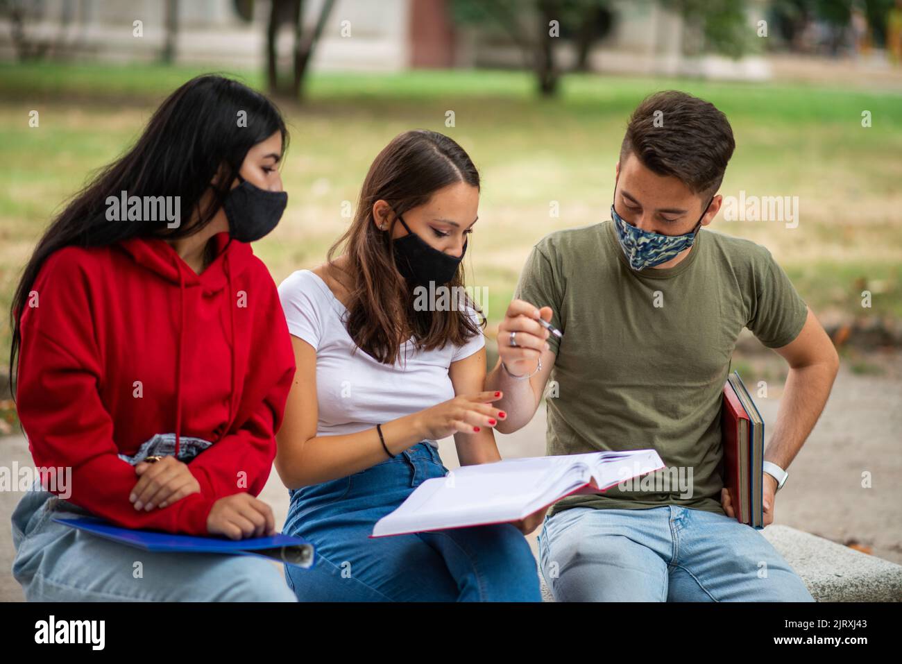 Three students studying together sitting on a bench outdoor and wearing ...