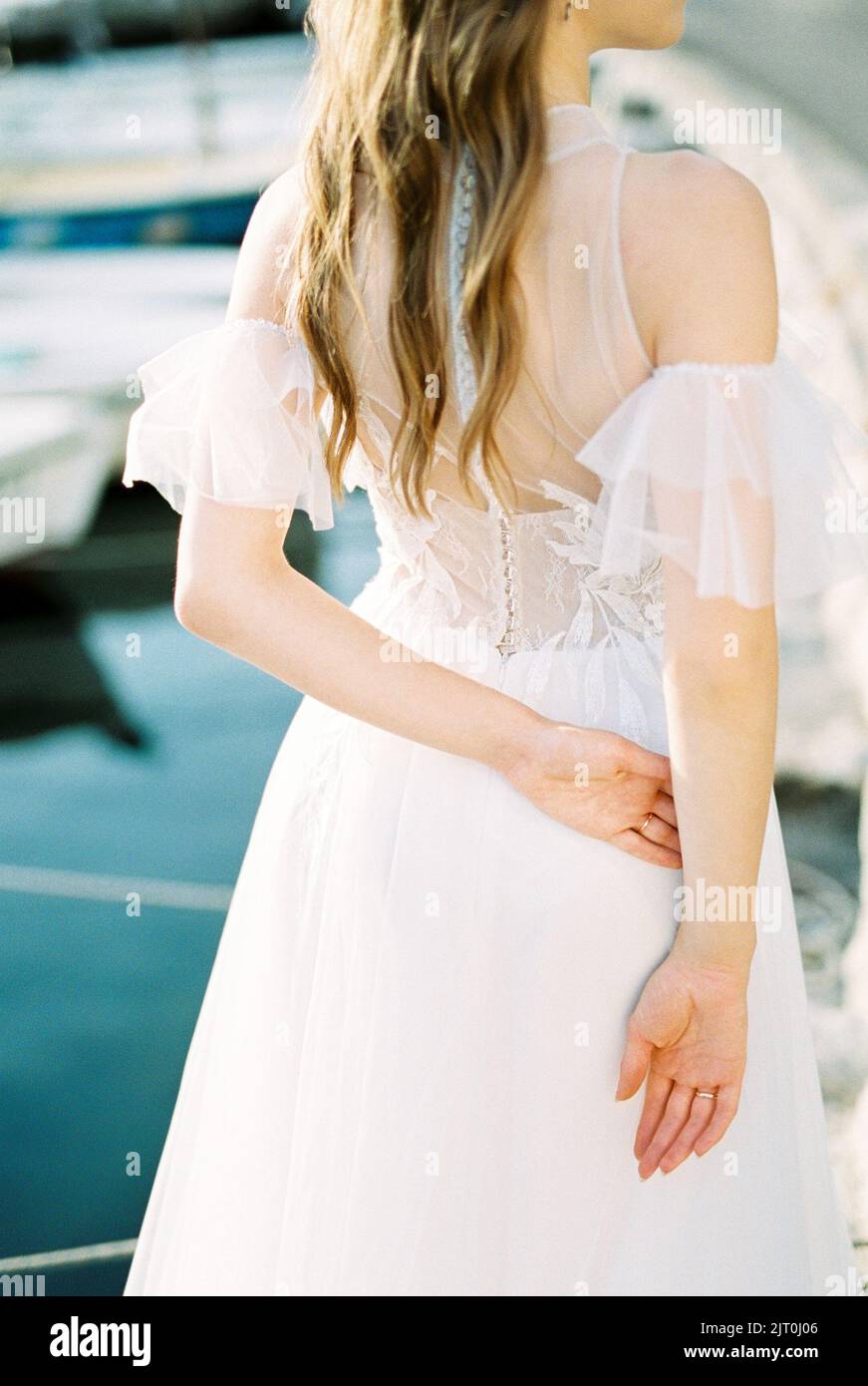 Bride in a white dress stands with her hands behind her back. Back view ...