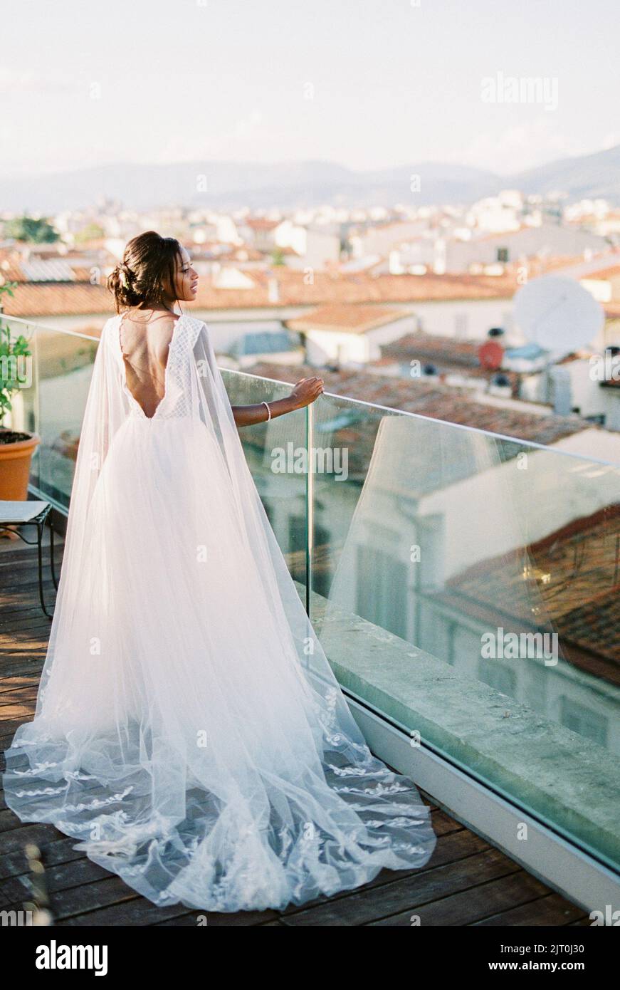 Bride stands near the glass railing and looks at the roofs of old ...