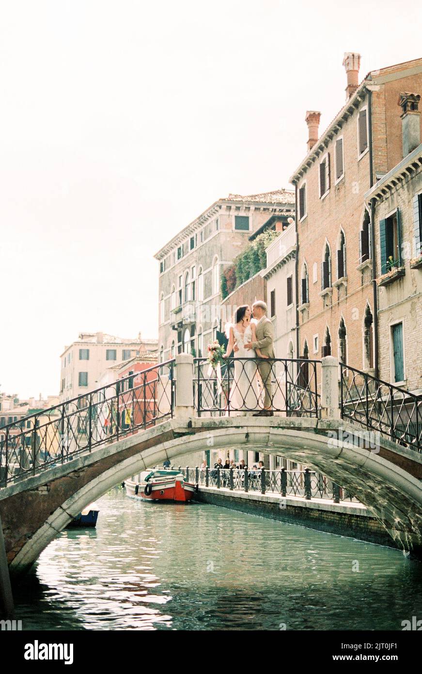 Bride and groom stand on a stone bridge over a canal against the ...