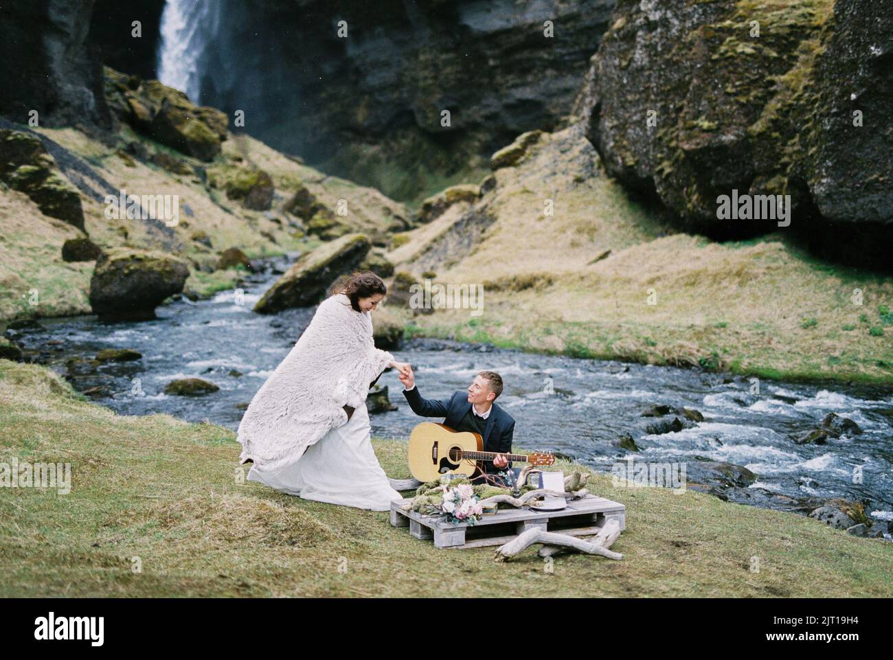 Groom sits with a guitar next to bride standing in a blanket on the ...