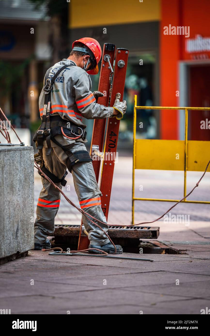 Brazilian maintenance worker holding a step ladder going under manhole ...