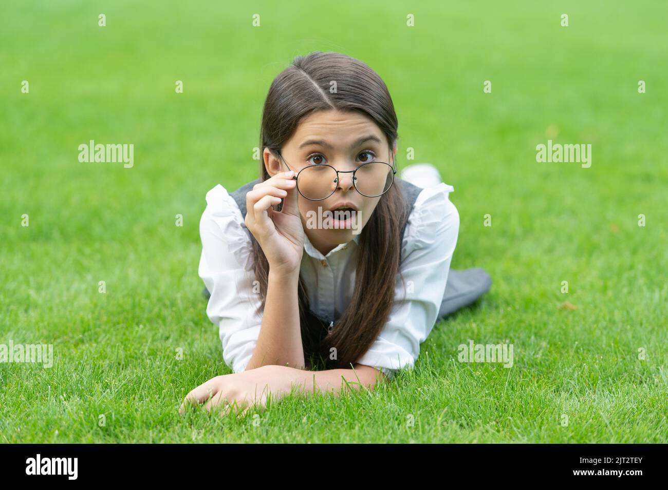 Portrait of surprised teen girl adjusting glasses lying on grass after ...