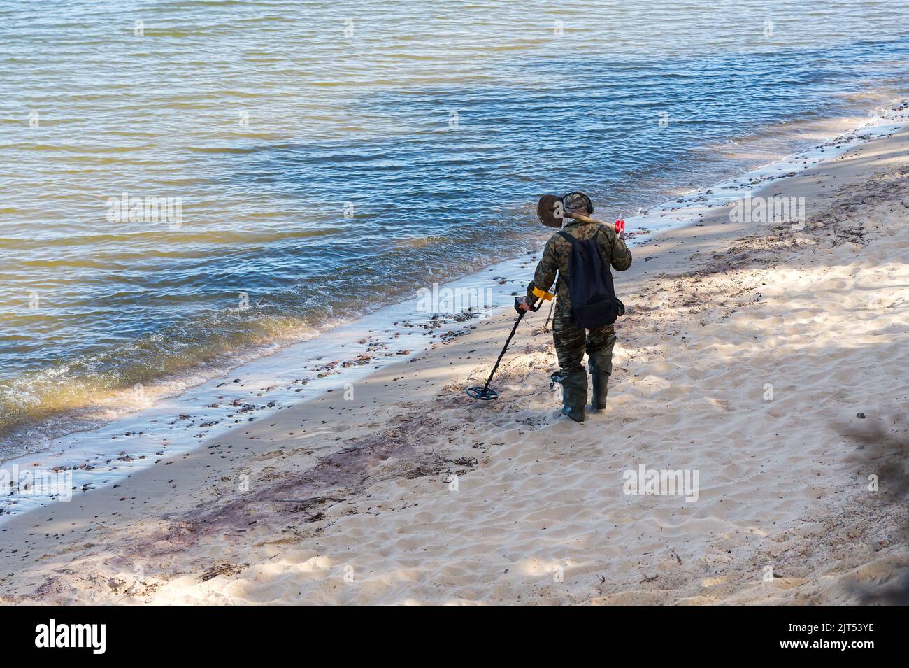 A man with a metal detector walks at the water's edge of a sandy beach ...