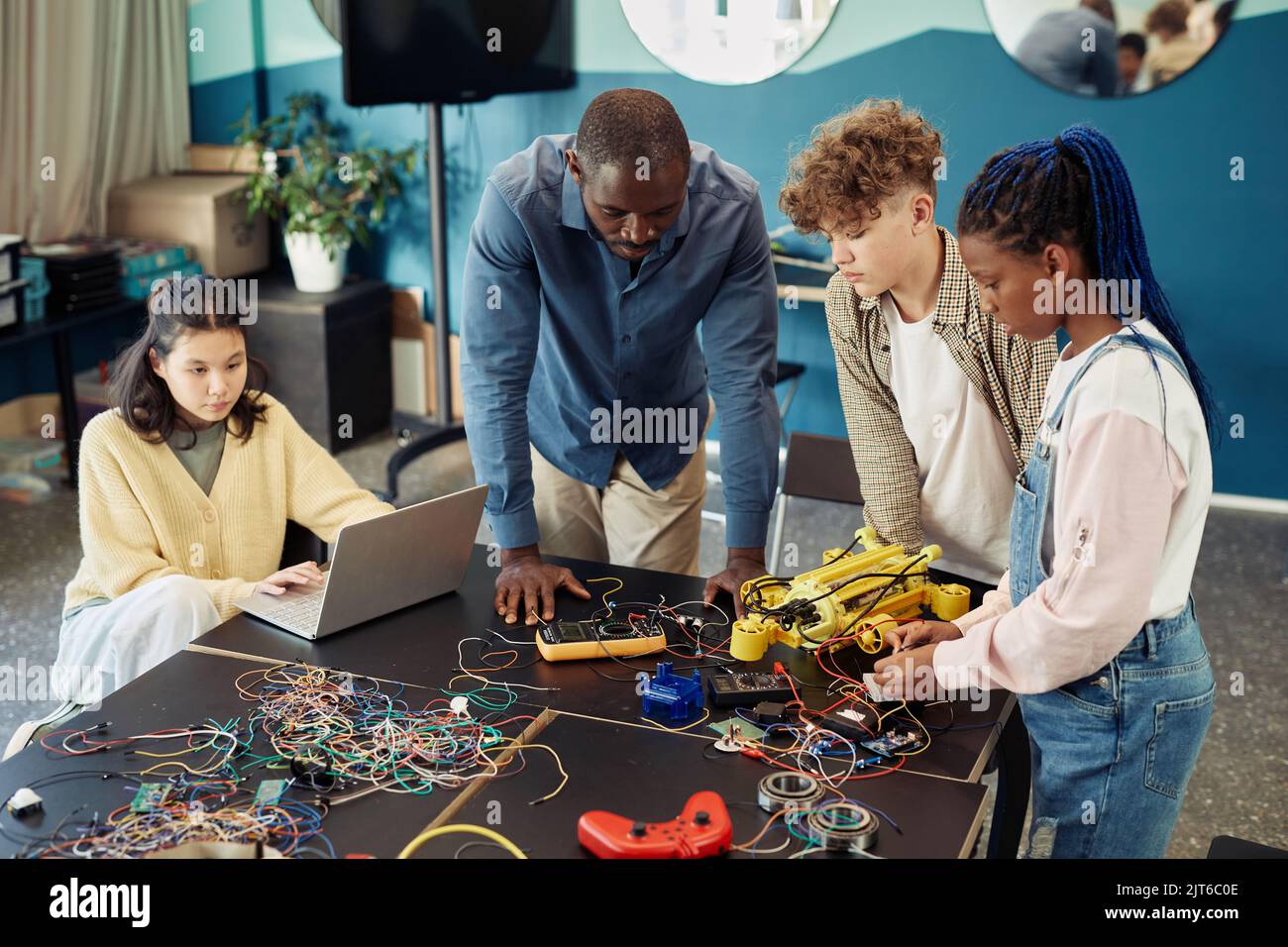 Portrait of male teacher with diverse group of kids building robots during engineering class at ...