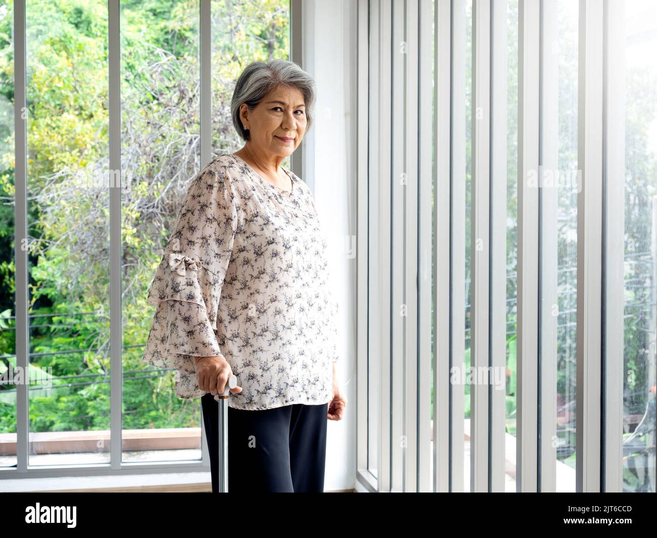 Asian senior woman, white hair standing with a cane near the glass ...