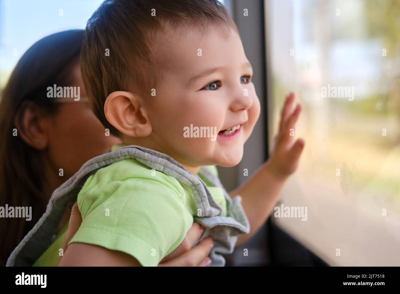 A happy toddler boy looks out the bus window while sitting in the arms ...