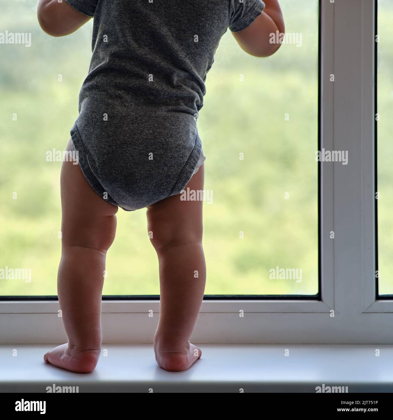 Toddler baby is standing by the window on the windowsill. Child at the ...