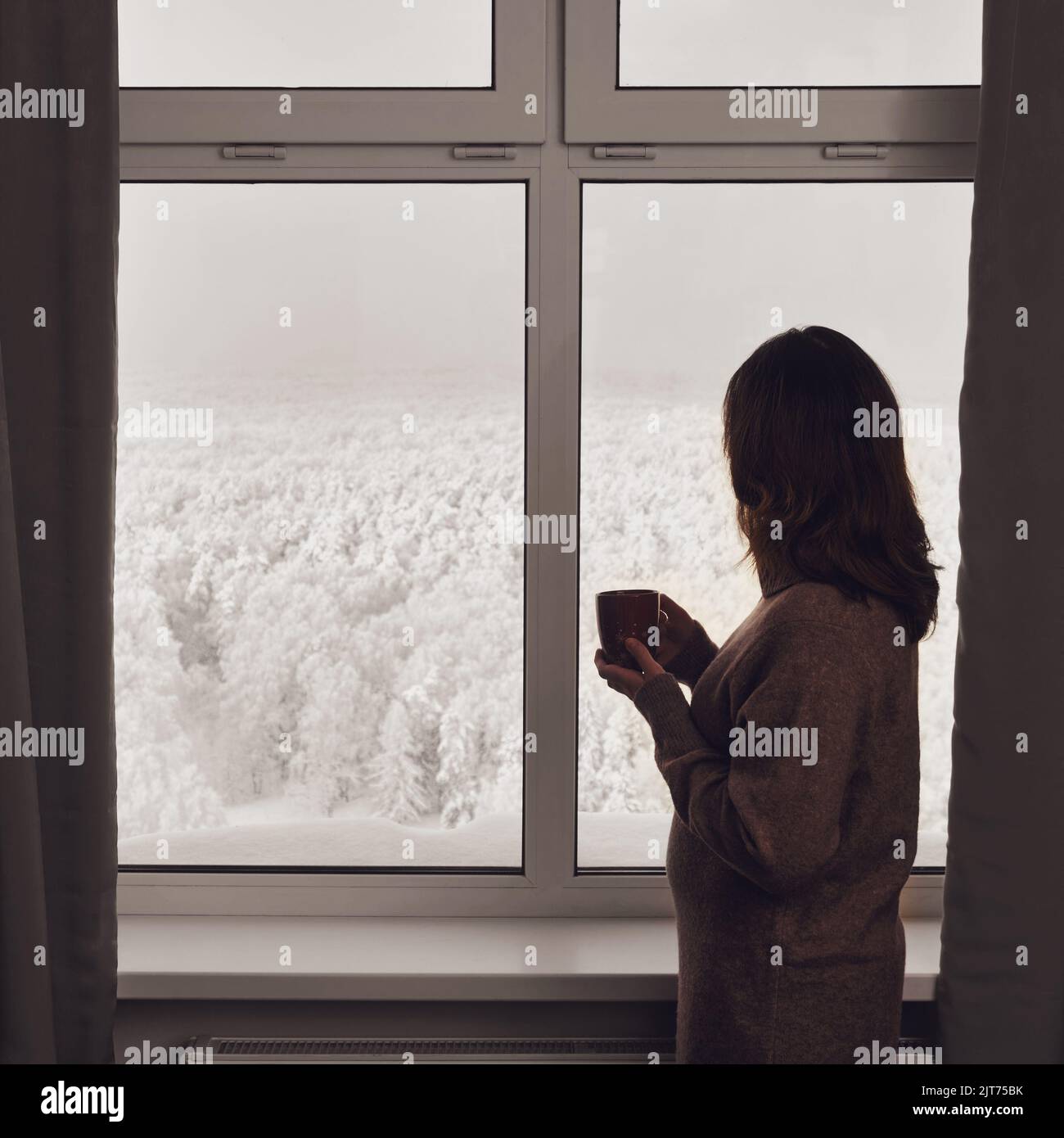 A woman with cup stands at a winter window with trees in the snow. A ...