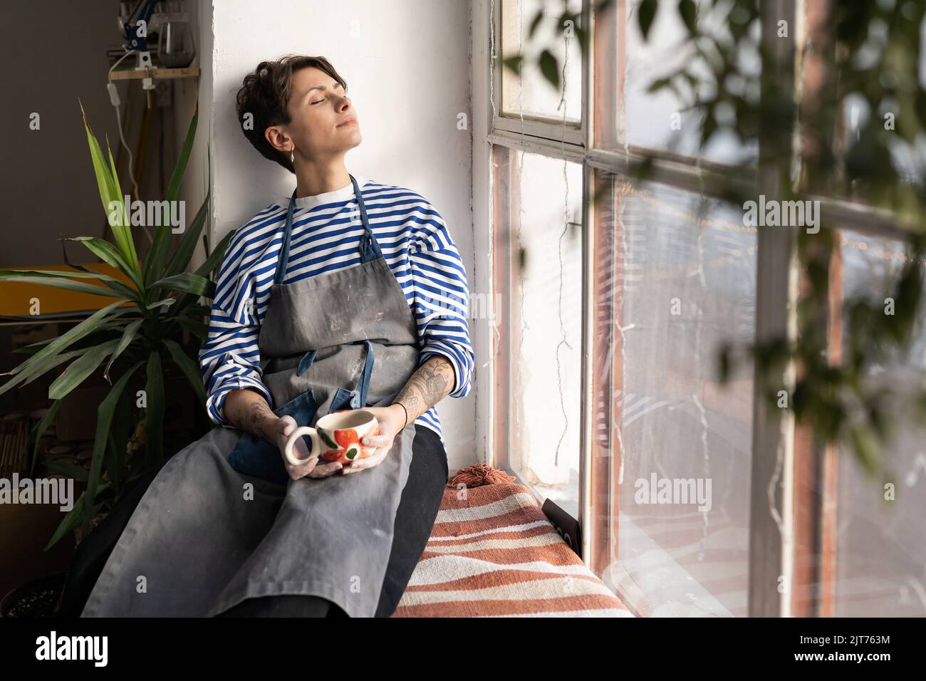 Female artisan holding cup of tea sitting on windowsill taking break ...