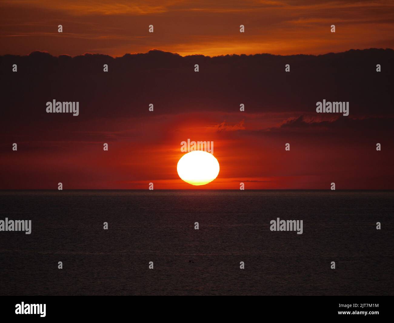 Deep red full sunset beneath a grey cloudbank over a dark sea ...
