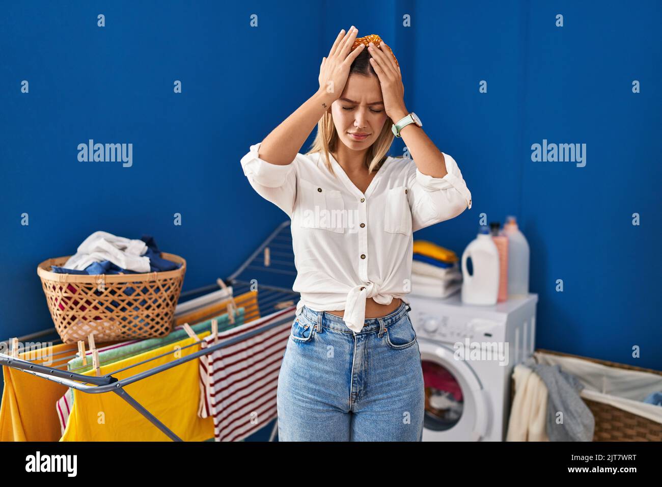 Young blonde woman at laundry room suffering from headache desperate ...