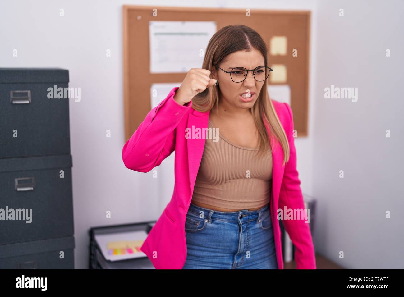 Young blonde woman standing at the office angry and mad raising fist ...