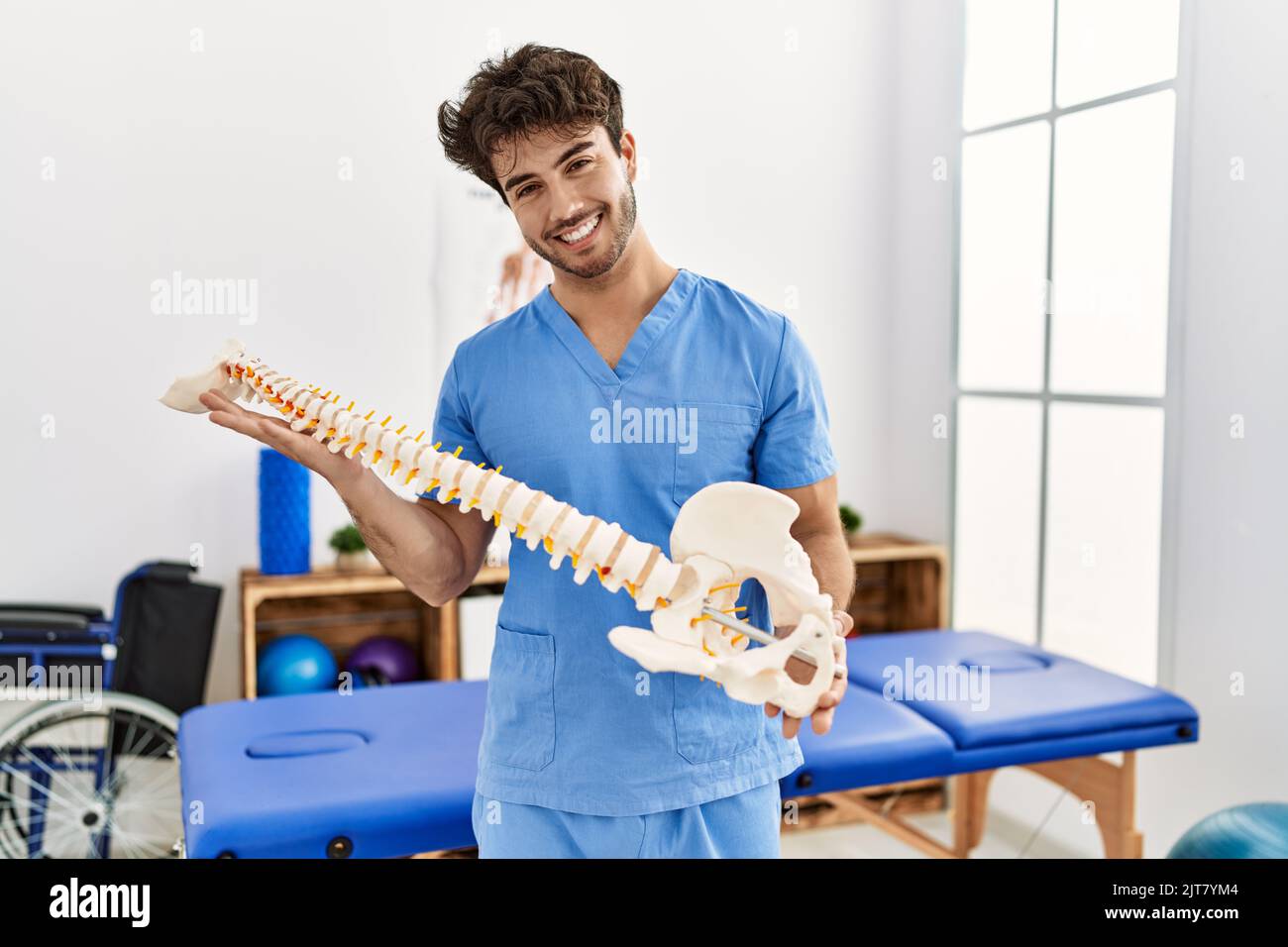 Young hispanic man wearing physio therapist uniform holding anatomical ...