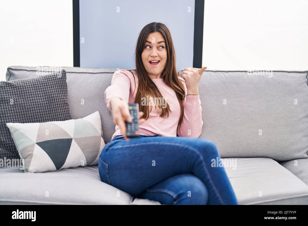 Young brunette woman holding television remote control pointing thumb ...