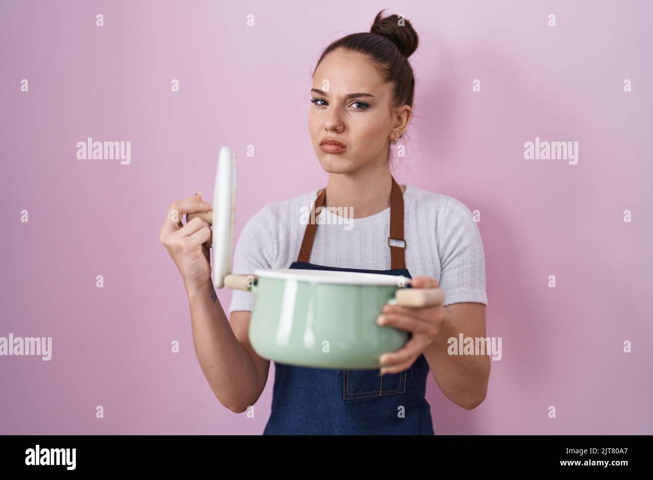Young hispanic girl wearing apron holding cooking pot skeptic and ...