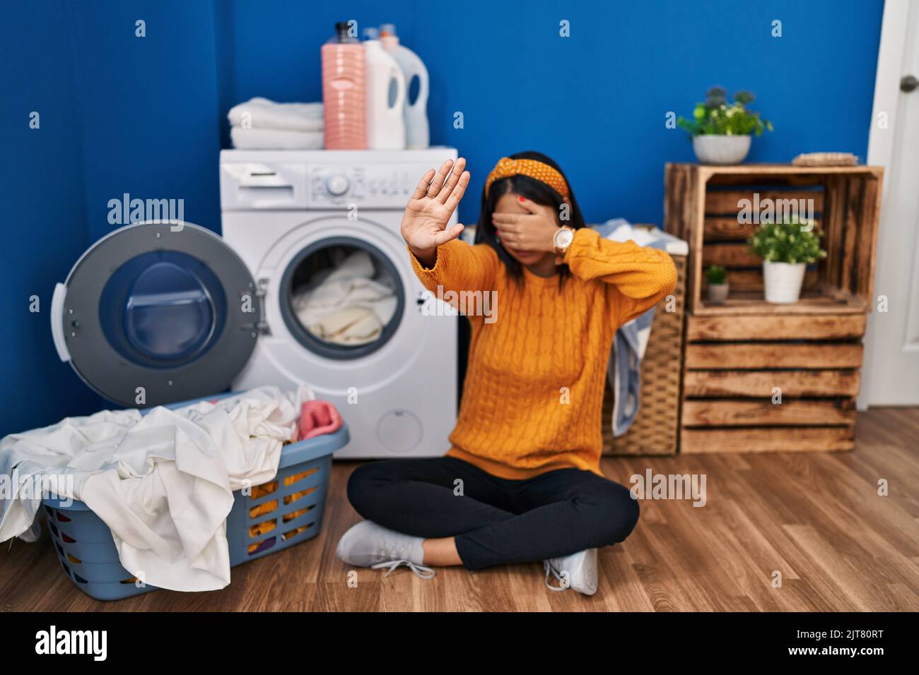 Young hispanic woman doing laundry covering eyes with hands and doing ...