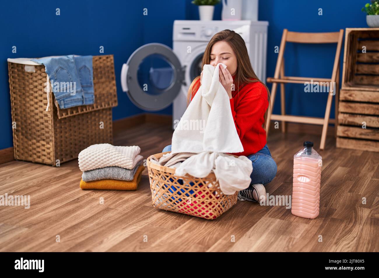 Young blonde woman smelling clothes sitting on floor at laundry room ...