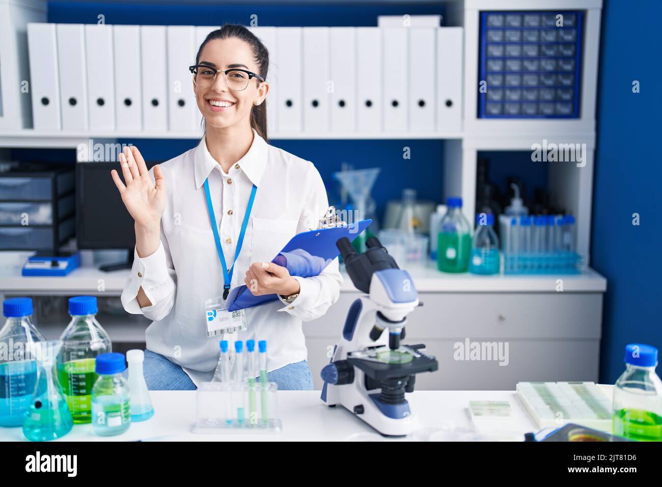 Young brunette woman working at scientist laboratory waiving saying ...